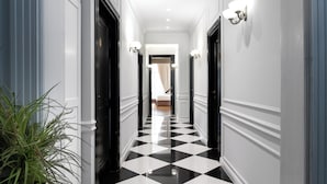 Um corredor com chão quadriculado em preto e branco, paredes brancas e portas pretas.