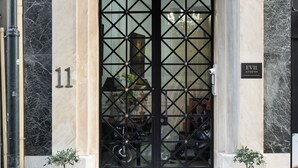 Entrada de uma residência de luxo neoclássica, com um grande portão preto, colunas e uma placa com a inscrição "EV11 ATHENS Neoclassical Luxury Residence".