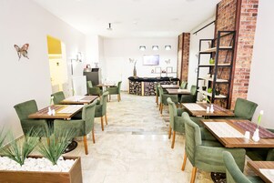 Uma área de refeições moderna com cadeiras estofadas em verde e mesas de madeira, uma parede de destaque em tijolo e uma mesa de recepção ao fundo.
