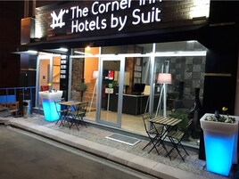 Fachada de hotel com letreiro de neon, mesas ao ar livre e um grande vaso de plantas.