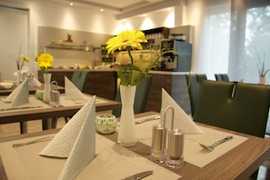 Uma mesa de jantar posta com centros geométricos, um vaso com flores amarelas e uma cozinha moderna ao fundo.