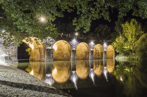 Uma ponte de pedra com arcos sobre águas calmas, rodeada por árvores e iluminada por postes de luz.