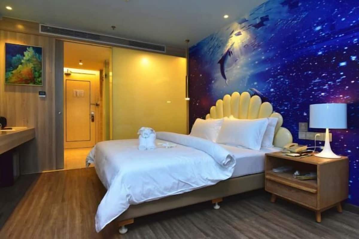 Quarto de hotel com cama, mesa de cabeceira, painel decorativo na parede e porta que dá para outro quarto.