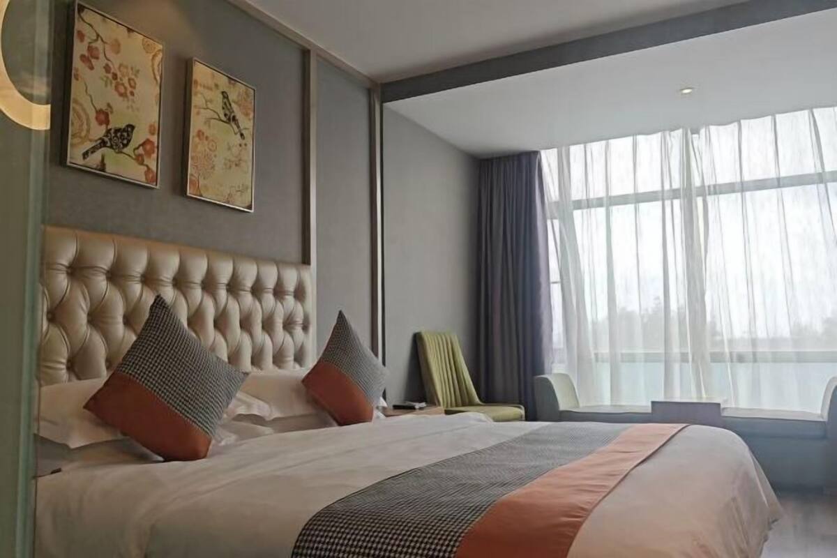 Quarto de hotel com uma cama grande, duas obras de arte na parede, uma janela com cortinas leves e um espaço de estar com uma cadeira e uma mesinha.