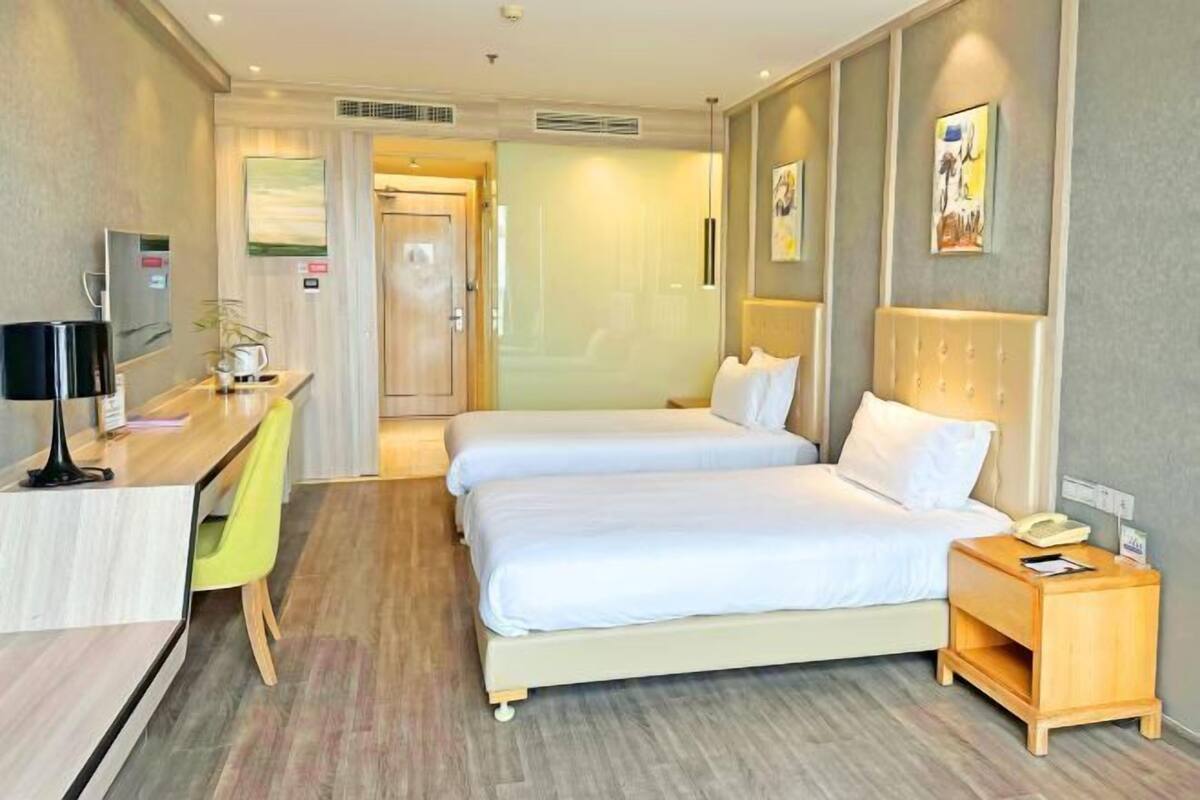 Quarto de hotel com duas camas, uma escrivaninha com cadeira, mesa de cabeceira com abajur e televisão.