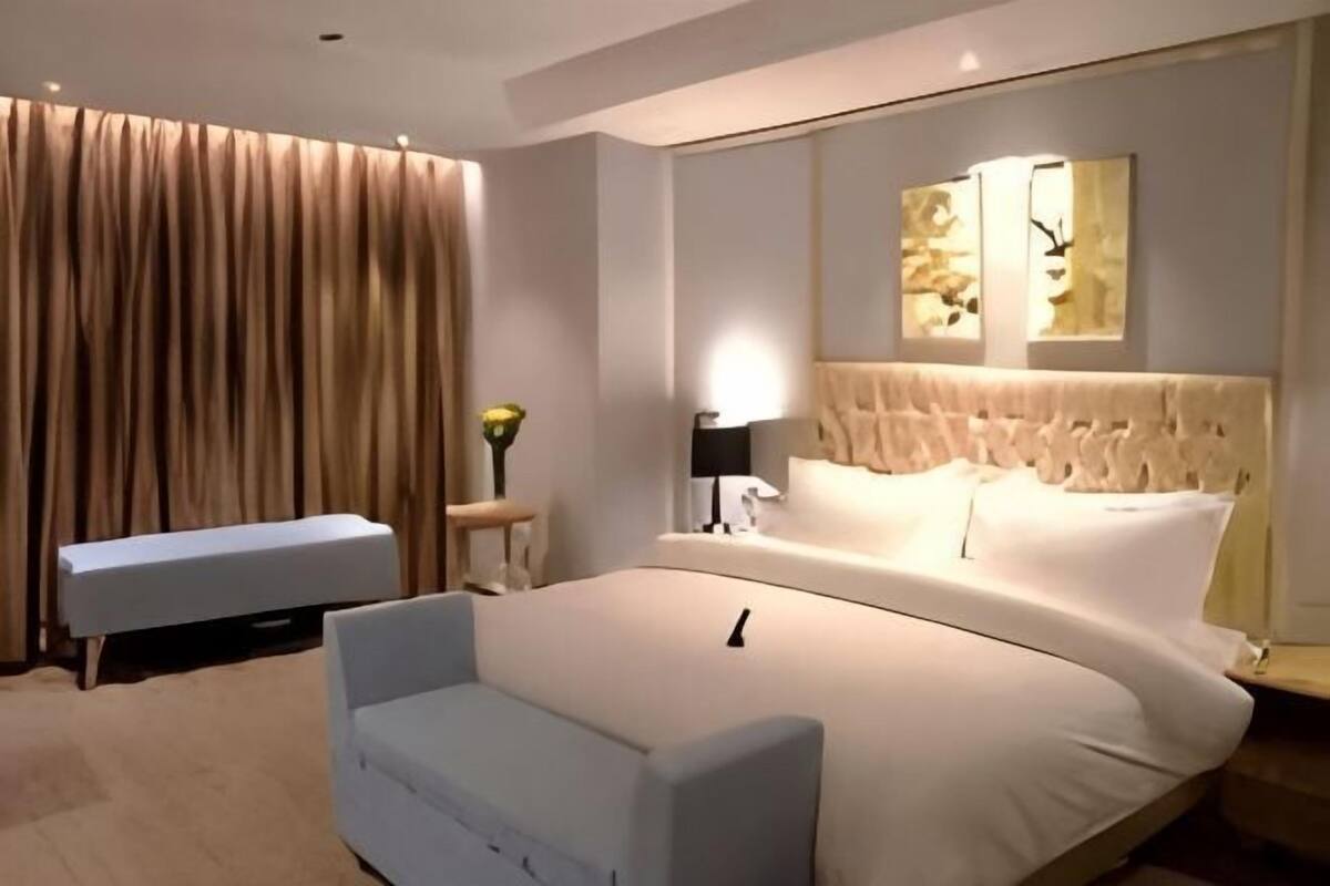 Quarto de hotel moderno com uma cama grande, uma mesa de cabeceira, um abajur e um banco.