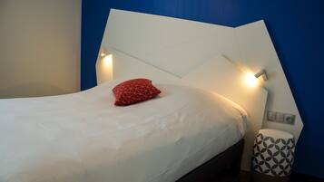 Cama bem arrumada com roupa de cama branca e um travesseiro geométrico vermelho, uma luminária de cabeceira e um banquinho estampado.