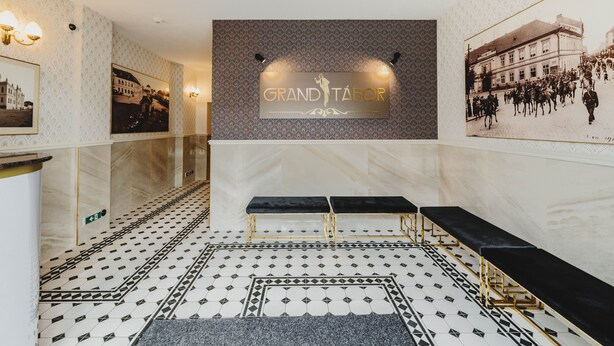 Um saguão de hotel com piso estampado, um banco e uma placa fixada na parede com a inscrição 'GRAND TÁBOR'.