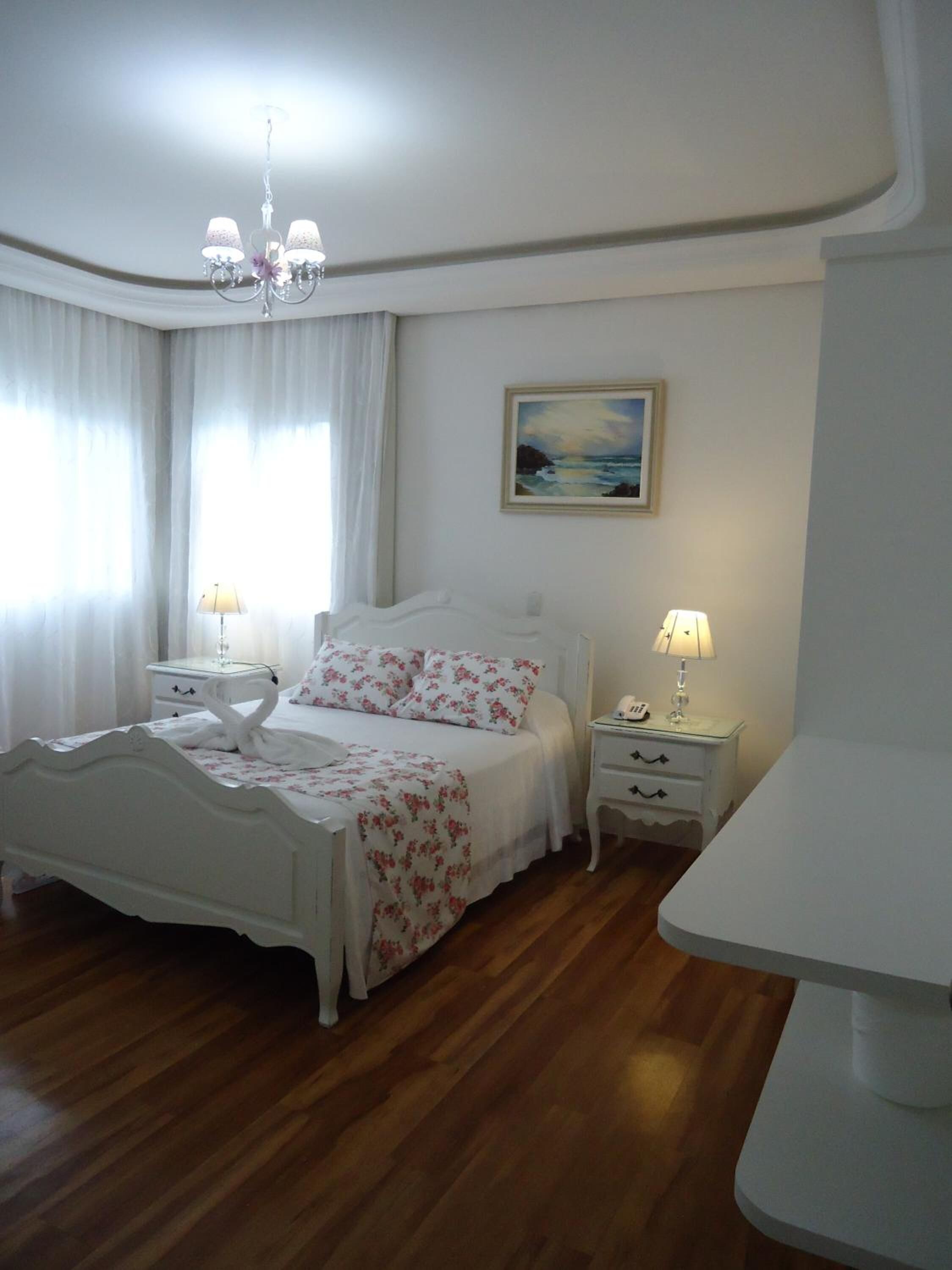 Quarto com cama, duas mesas de cabeceira, um candelabro, um quadro e uma janela com cortinas.