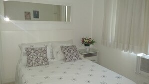 Quarto com cama, duas almofadas, mesa de cabeceira com um vaso de flores, espelho na parede e janela com cortinas.