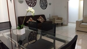 Uma sala de estar moderna com uma mesa de jantar de vidro, um sofá e duas poltronas. Há obras de arte decorativas nas paredes e um vaso com planta.