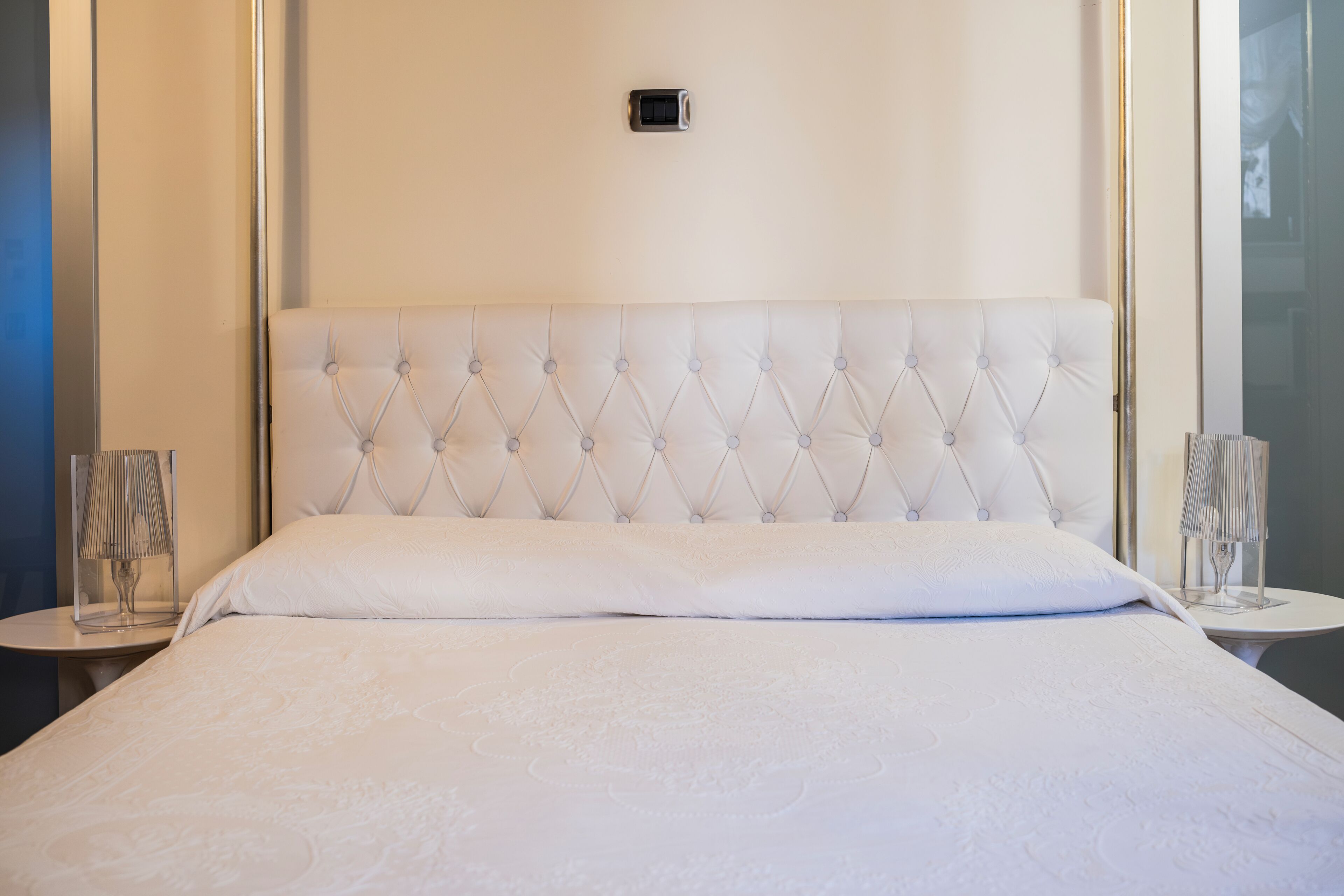 Uma cama bem arrumada com cabeceira estofada, duas mesas de cabeceira com abajures e um pequeno painel de controle fixado na parede.