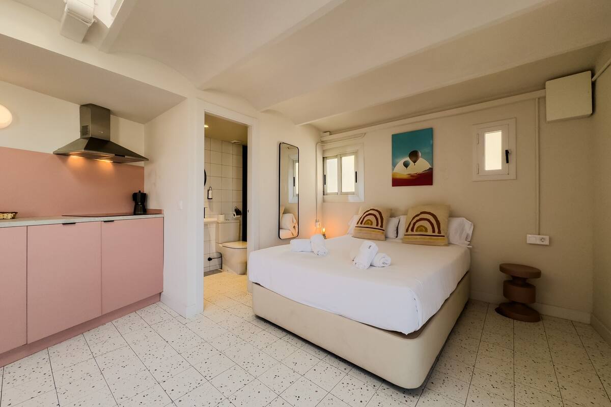 Quarto com cama, mesa de cabeceira, janela e banheiro visível por uma porta aberta.