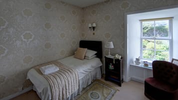 Quarto com cama, mesa de cabeceira, abajur e janela com vista para o verde.