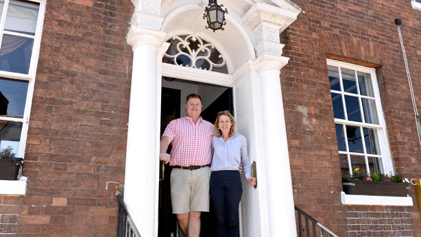 Um homem e uma mulher em frente à entrada de um edifício de tijolos, com um arco branco e um candeeiro preto acima.