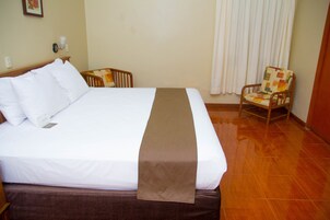 Uma cama bem arrumada com um espalha cama castanho, uma cadeira de madeira com uma almofada colorida e uma cortina branca numa janela.