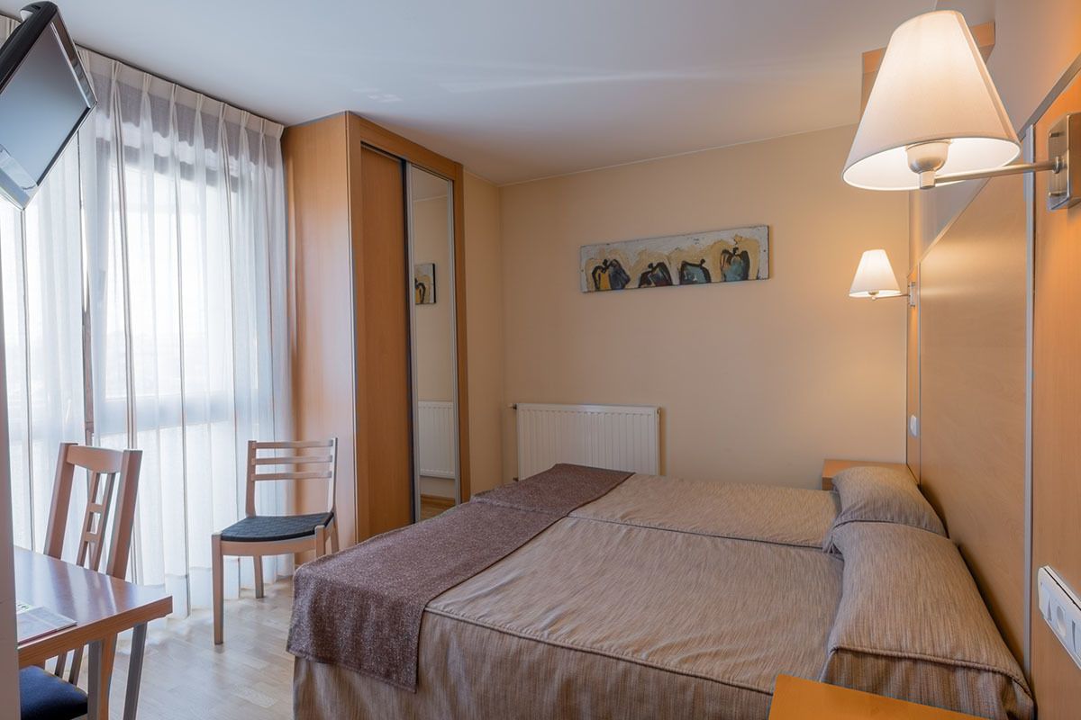 Quarto de hotel com cama, uma cadeira, escrivaninha e luminária.