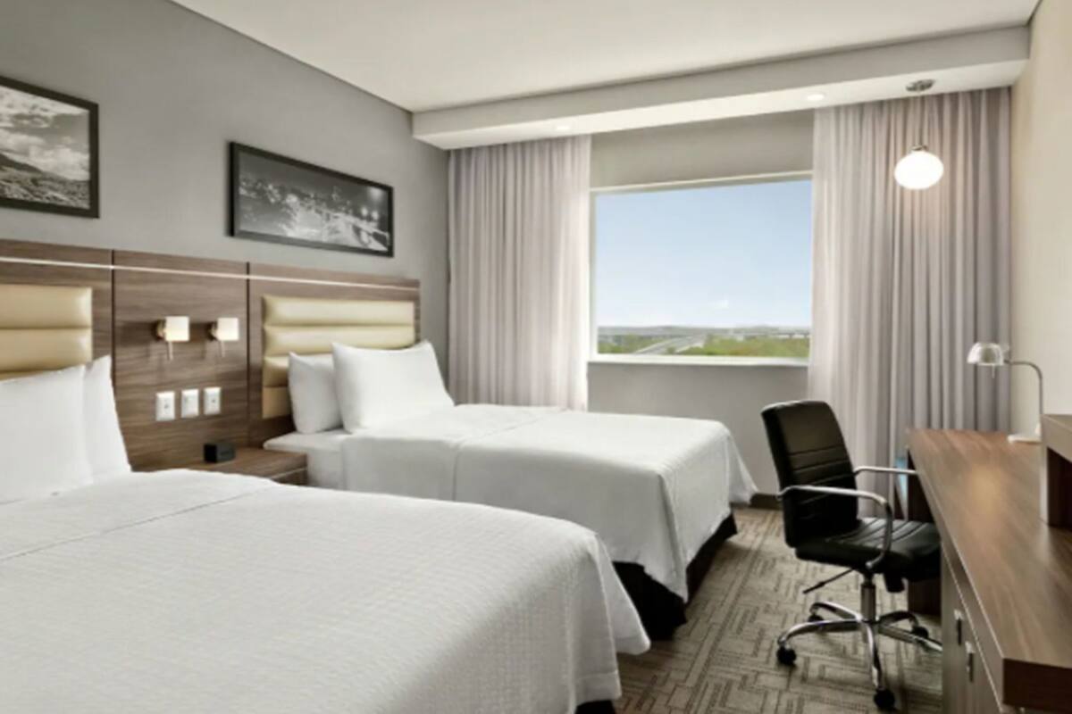 Quarto de hotel com duas camas, uma escrivaninha, uma cadeira, uma janela com vista e uma luminária.