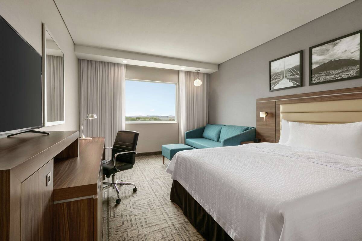 Quarto de hotel com uma cama grande, uma escrivaninha com cadeira, um sofá, uma TV e uma janela com cortinas.