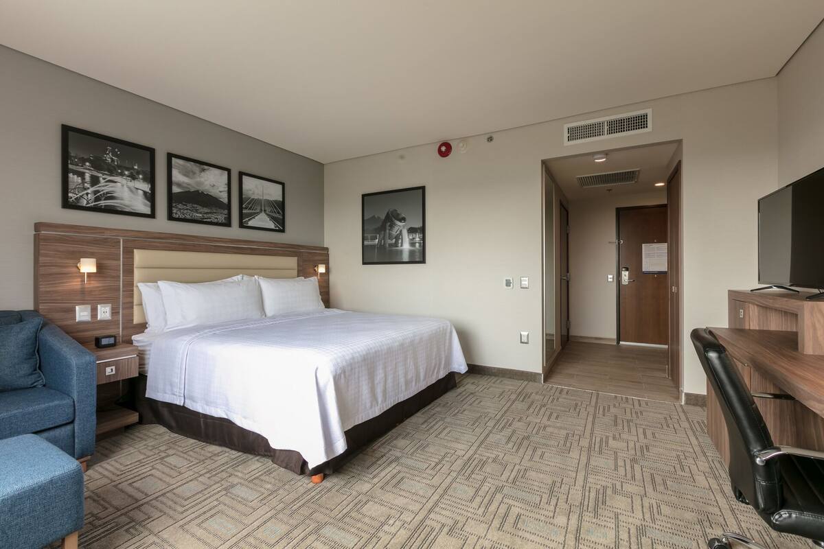 Quarto de hotel com uma cama grande, uma escrivaninha com cadeira, televisão e obras de arte nas paredes.