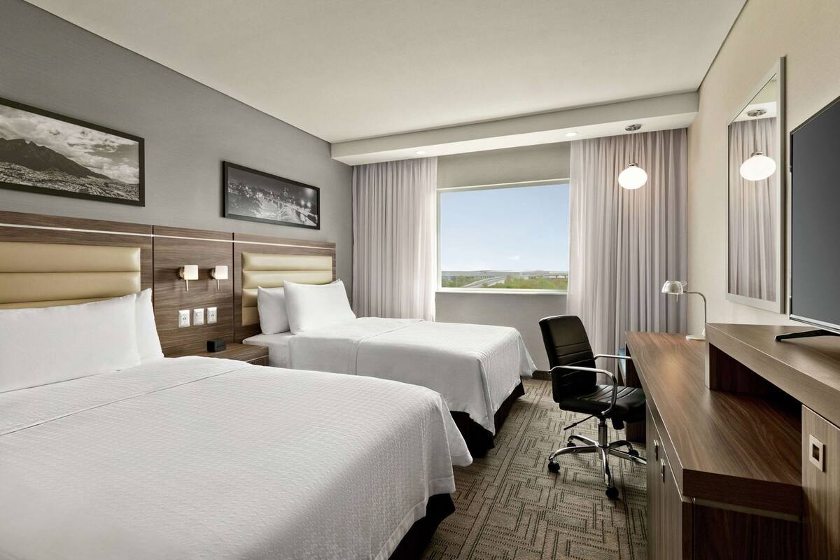 Quarto de hotel com duas camas, uma escrivaninha, uma cadeira, uma televisão e uma janela com vista.