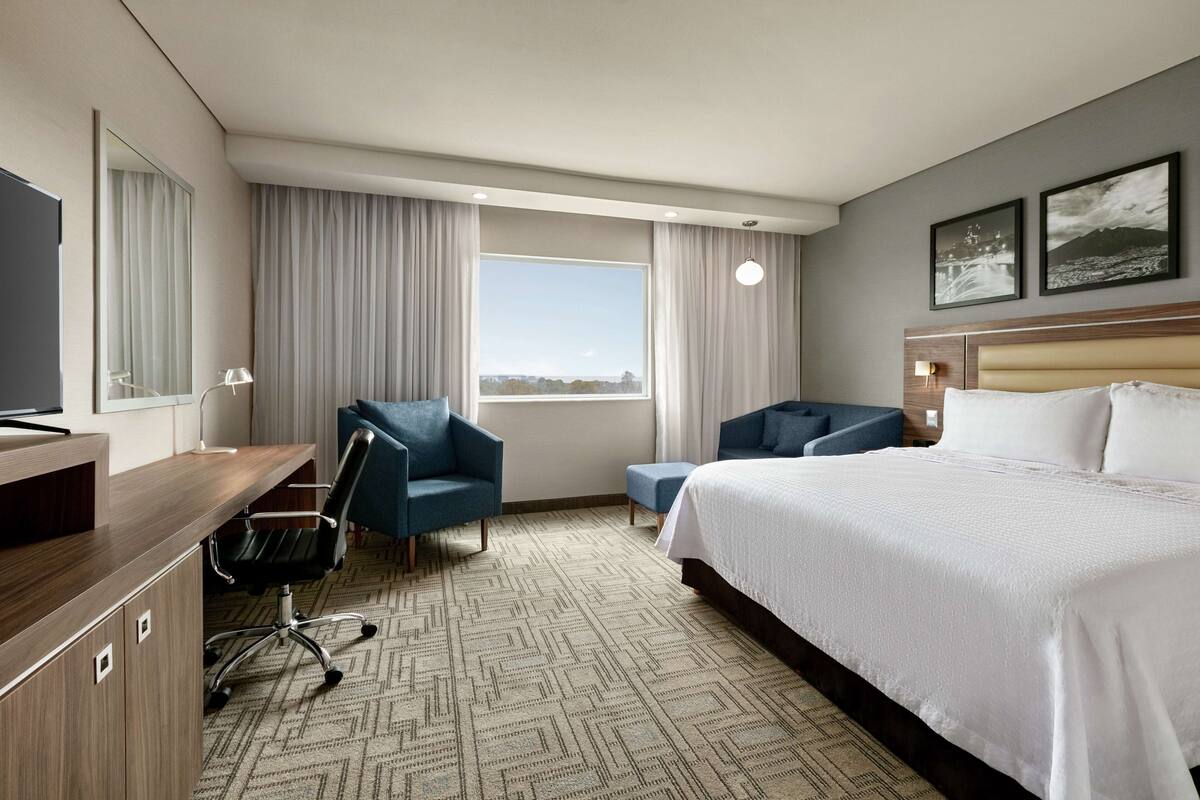 Quarto de hotel com cama grande, escrivaninha, duas cadeiras, televisão e janela com cortinas.