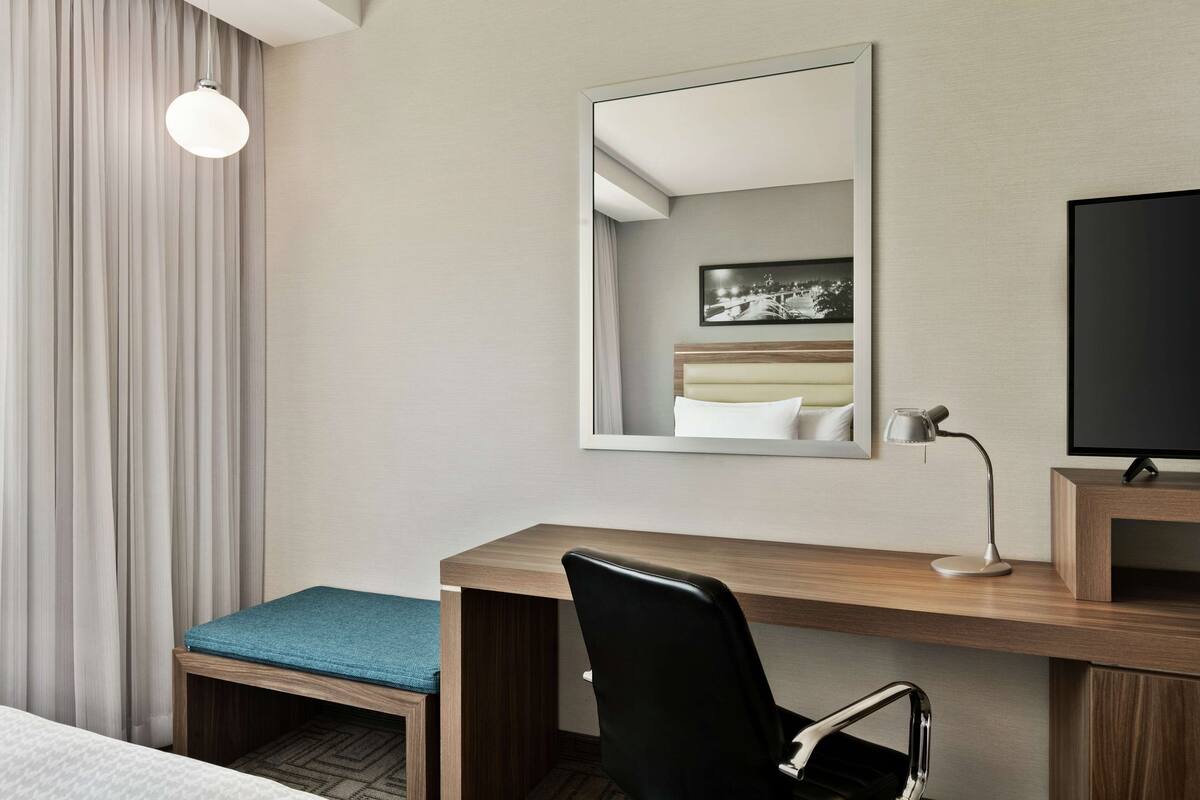 Quarto de hotel com cama, escrivaninha com cadeira, televisão, espelho e um quadro na parede.