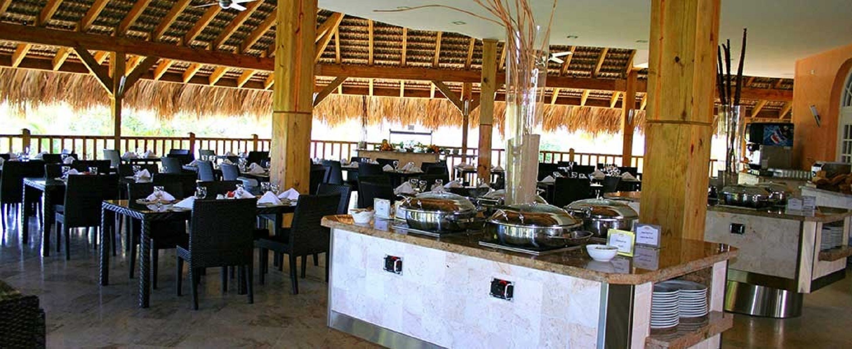 Restaurante em estilo buffet com várias mesas e cadeiras, um balcão central de serviço com recipientes de comida e uma estrutura de telhado de palha.