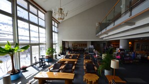 Um espaço amplo e interno, com grandes janelas, mesas e cadeiras de madeira, e diversas plantas.