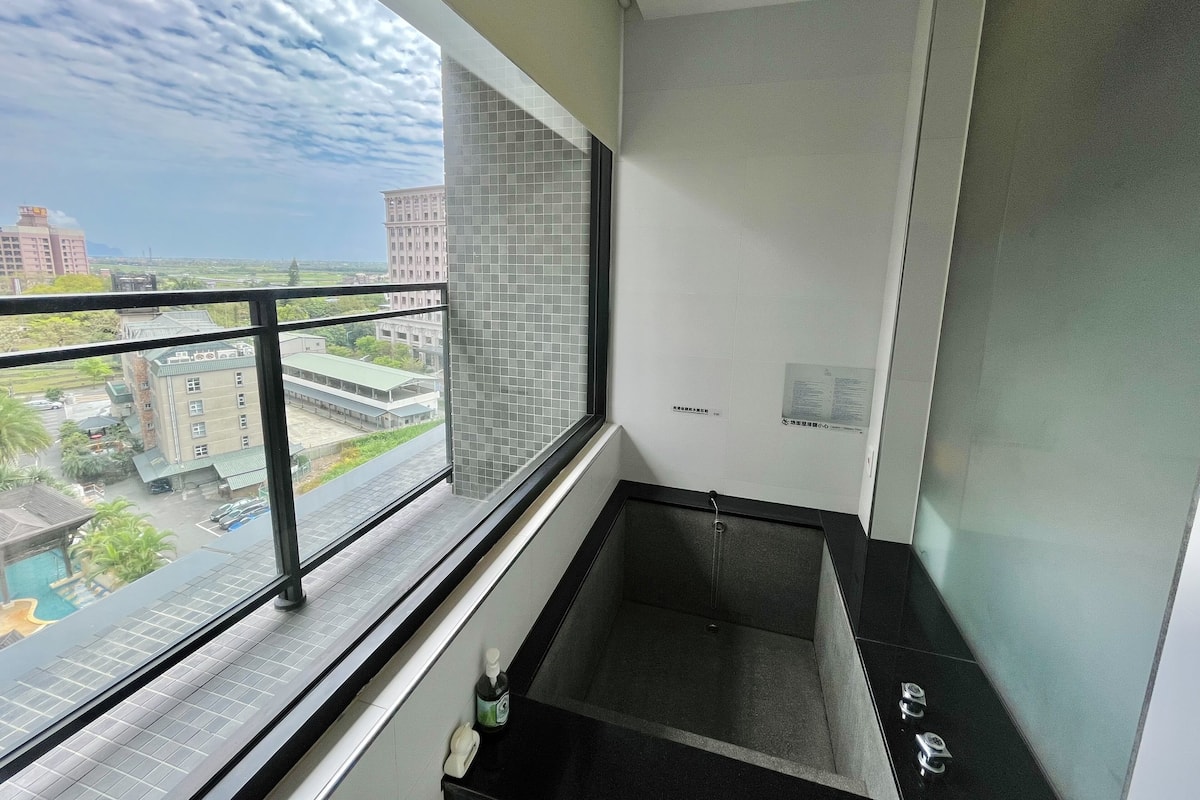 Um banheiro moderno com uma janela ampla, uma banheira e vista para edifícios e vegetação lá fora.
