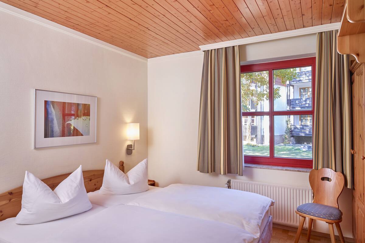 Quarto de hotel com teto de madeira, cama com roupa branca, uma cadeira, janela com cortinas e luminária fixada na parede.