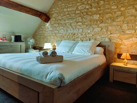 Quarto com parede de pedra, estrutura de cama de madeira, mesa de cabeceira com luminária e criado-mudo com abajur.