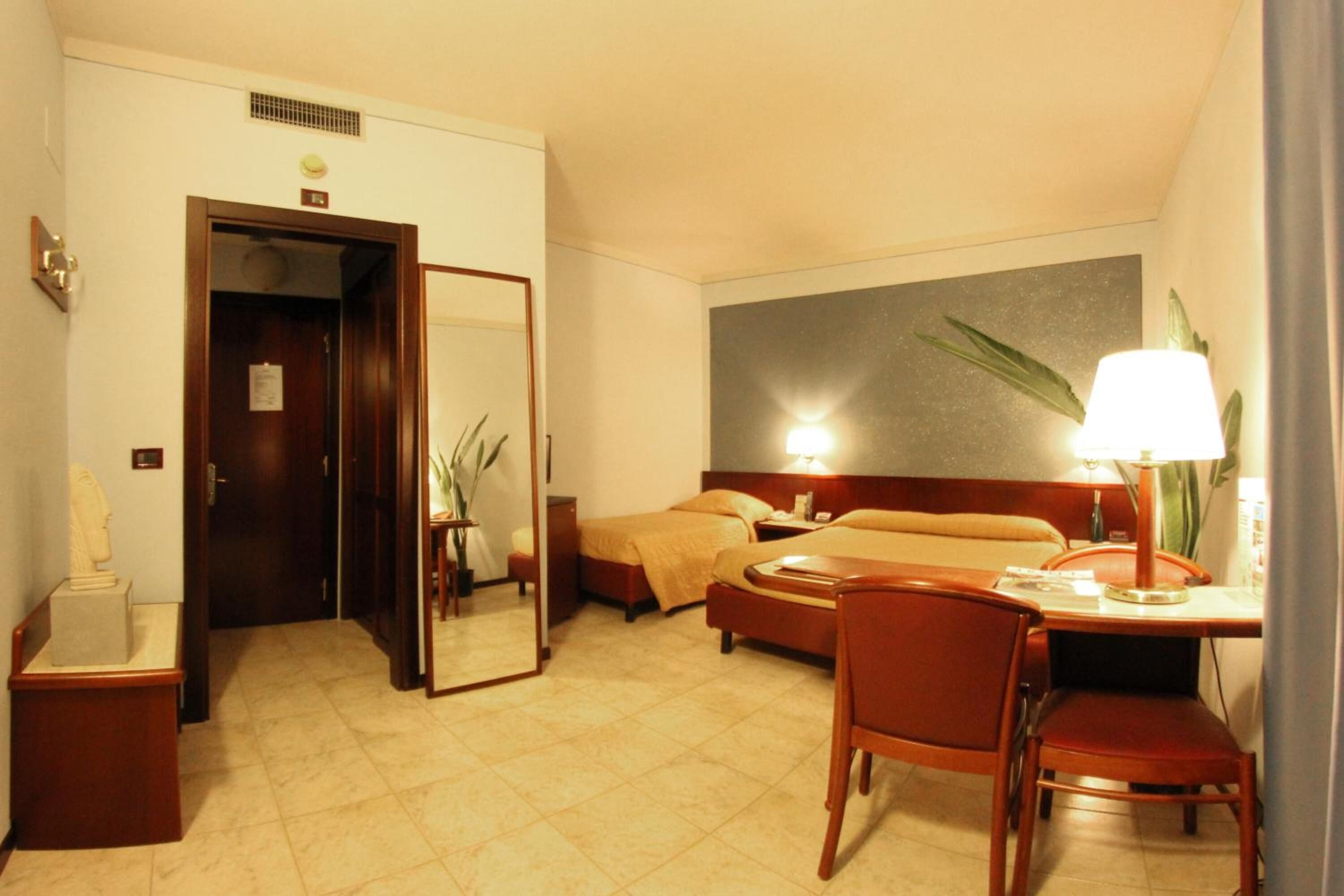 Quarto de hotel com uma cama, uma escrivaninha com cadeira, um espelho e uma luminária.