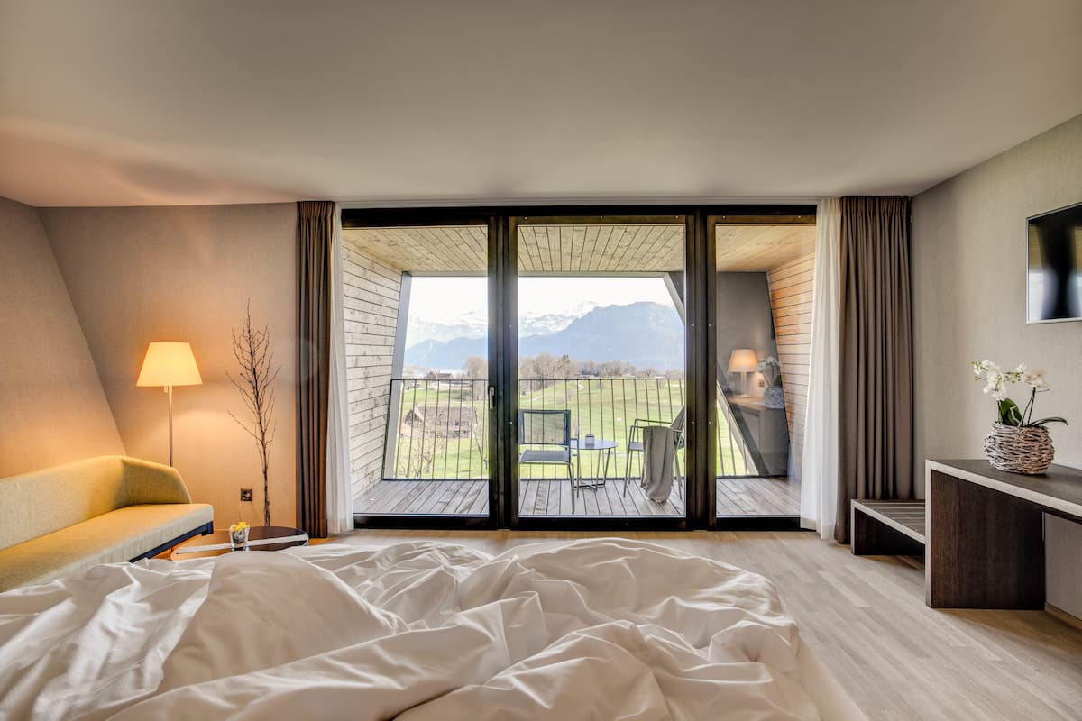 Quarto de hotel com uma cama grande, um sofá, uma mesa e vista para uma paisagem montanhosa através de uma porta deslizante.