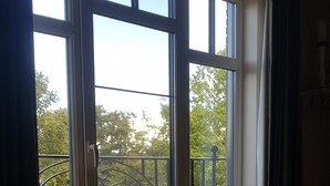 Quarto com uma grande janela, piso de madeira e guarda-corpo de metal na parte externa.
