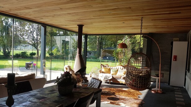 Um interior moderno com uma grande janela de vidro, teto de madeira, mesa de jantar com cadeiras, sofá, luminária pendente e uma gaiola decorativa.