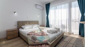Quarto moderno com cama grande, mesa de cabeceira, ar condicionado e vista para o exterior através de cortinas leves.