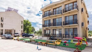 Um hotel com vários andares, varandas, área de recreação infantil com brinquedos e assentos, e estacionamento com carros.