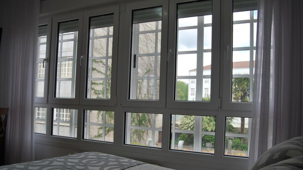 Quarto com uma grande janela, cama com colcha estampada e vista para o verde lá fora.