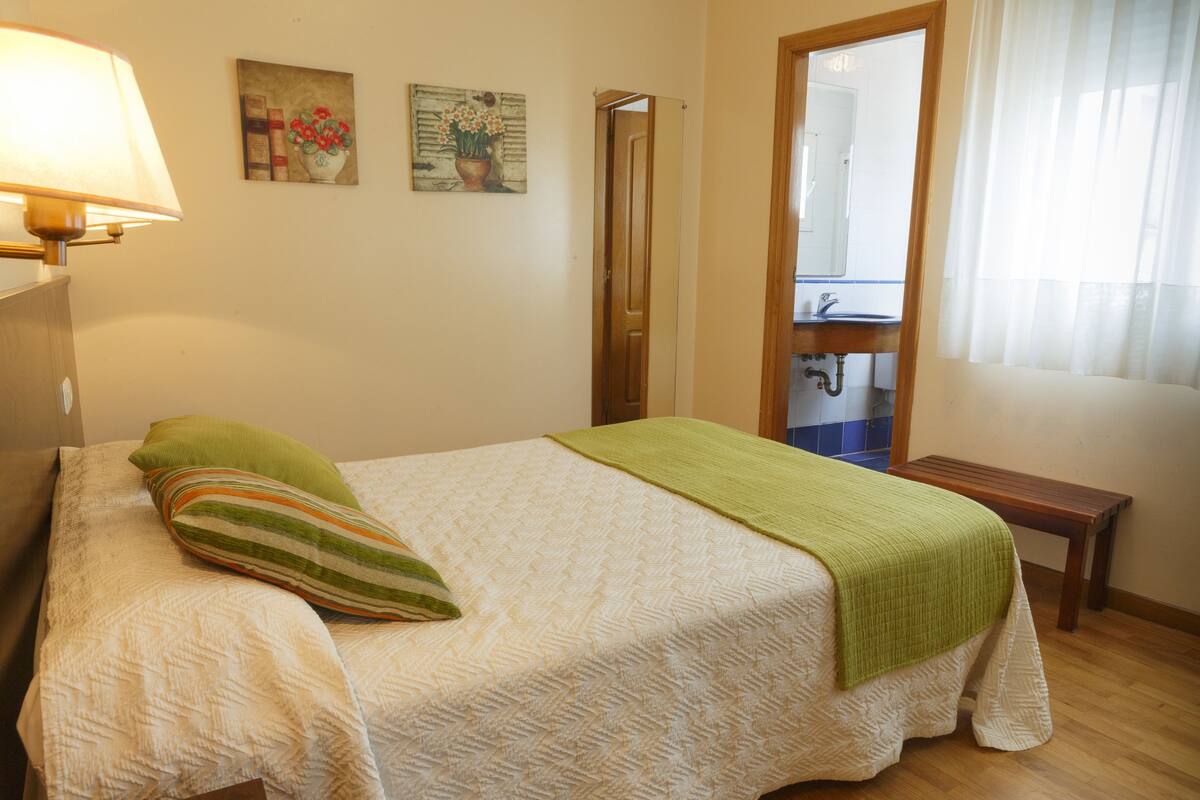 Cama bem arrumada com manta verde e travesseiro listrado, um banco de madeira, uma luminária na parede e um espelho refletindo um banheiro com pia e azulejos azuis.