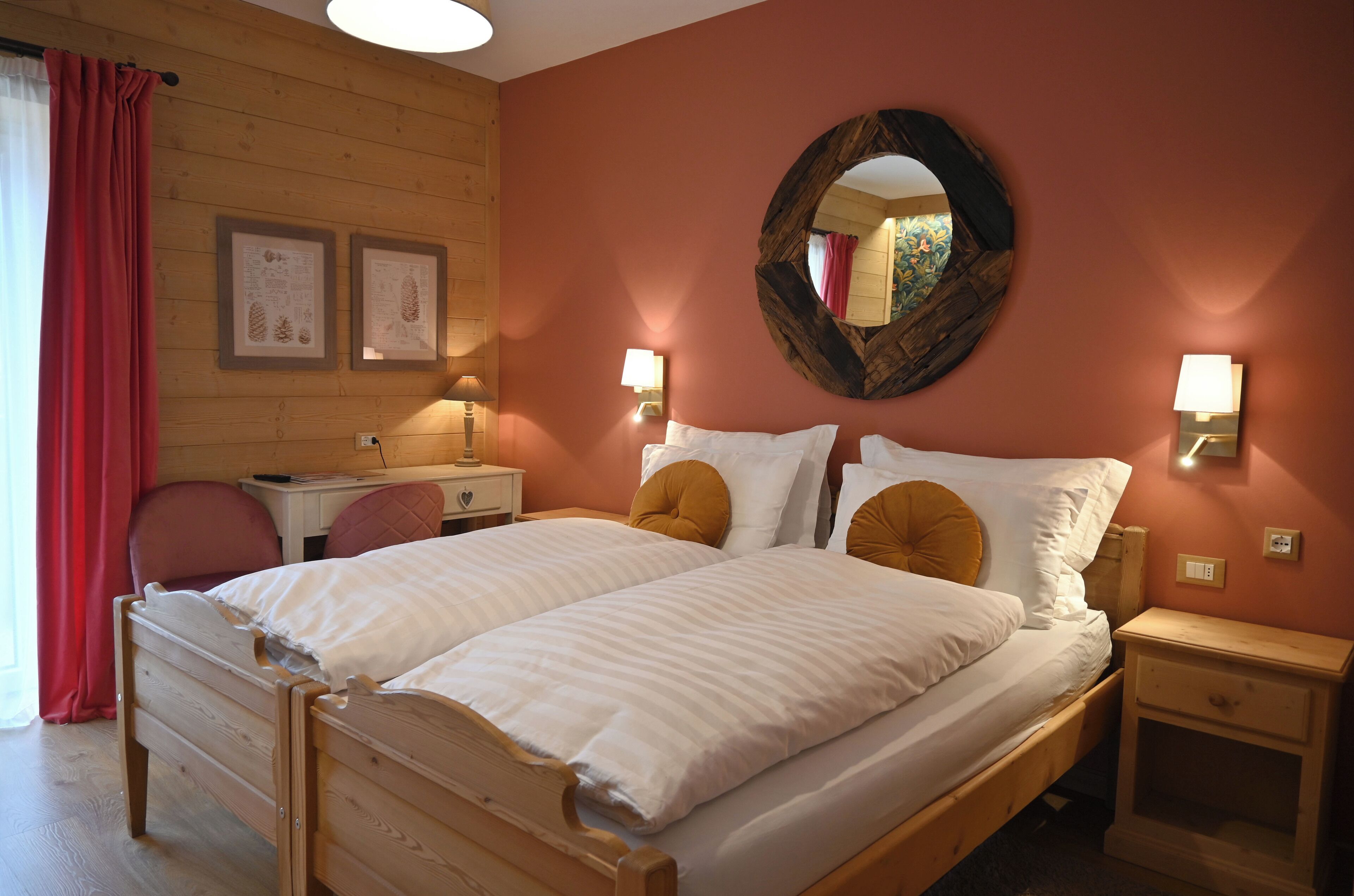 Uma cama de casal com roupa de cama branca, duas mesas de cabeceira com abajures, uma cabeceira de madeira, um espelho redondo grande e impressões botânicas emolduradas na parede.
