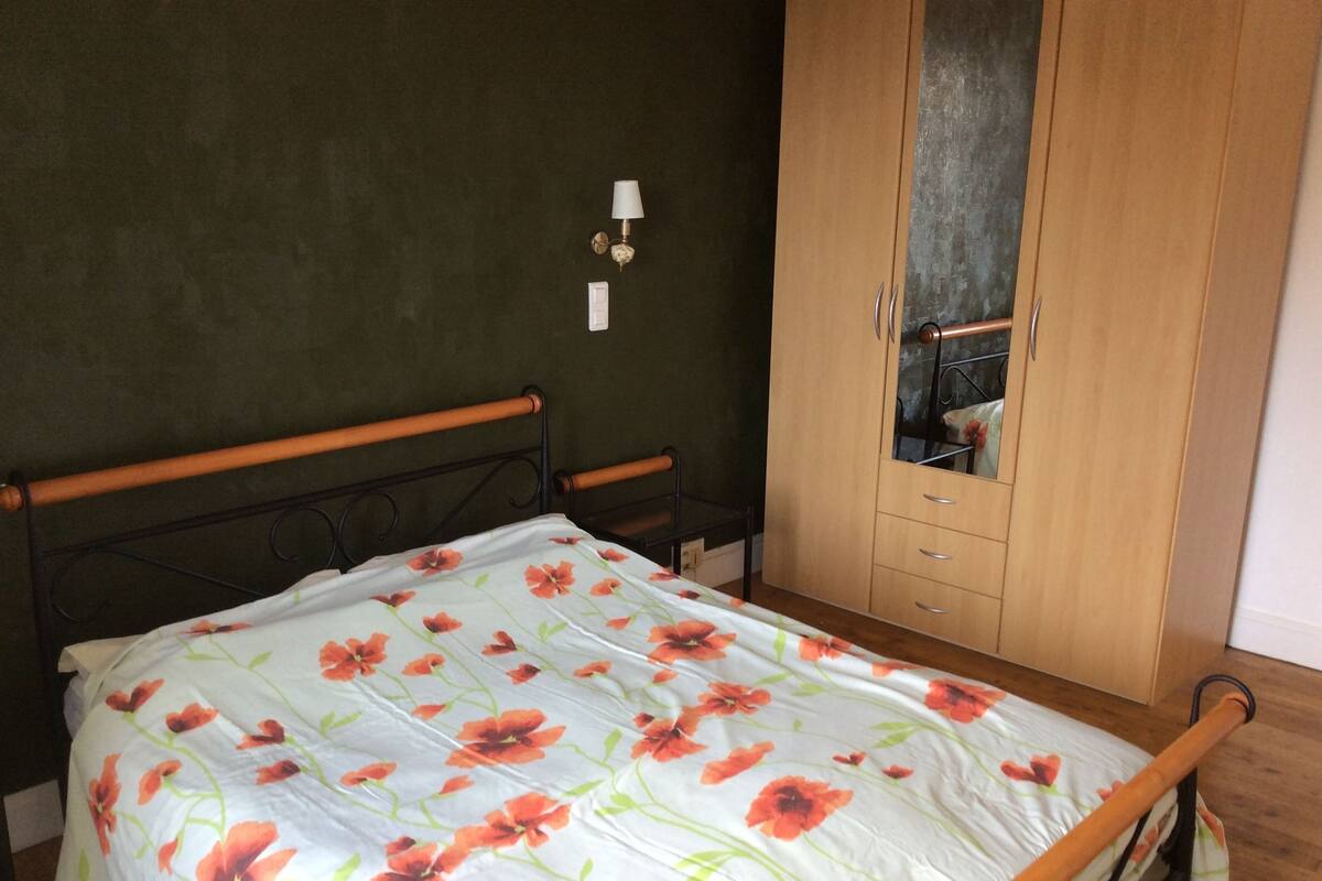 Um quarto com uma cama, um guarda-roupa de madeira, uma mesa de cabeceira e um abajur fixado na parede.