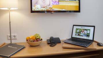 Quarto de hotel com secretária contendo um laptop, uma tigela de fruta, um telefone e uma televisão exibindo um noticiário.