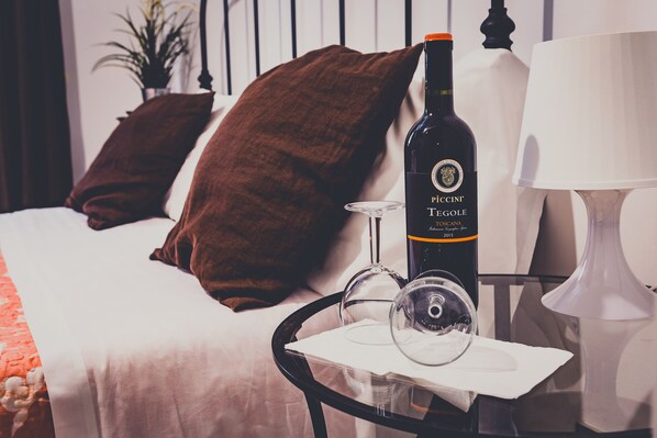 Cama arrumada com um edredom branco e castanho, uma garrafa de vinho, duas taças de vinho e uma pequena bandeja com um guardanapo sobre uma mesa de cabeceira.