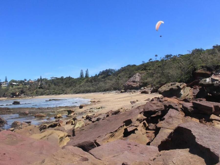 Uma praia rochosa com céu claro e um parapente ao longe.