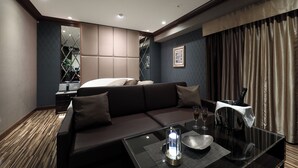 Quarto de hotel moderno com um sofá marrom escuro, uma mesa de centro de vidro, um minibar e uma janela grande com cortinas.