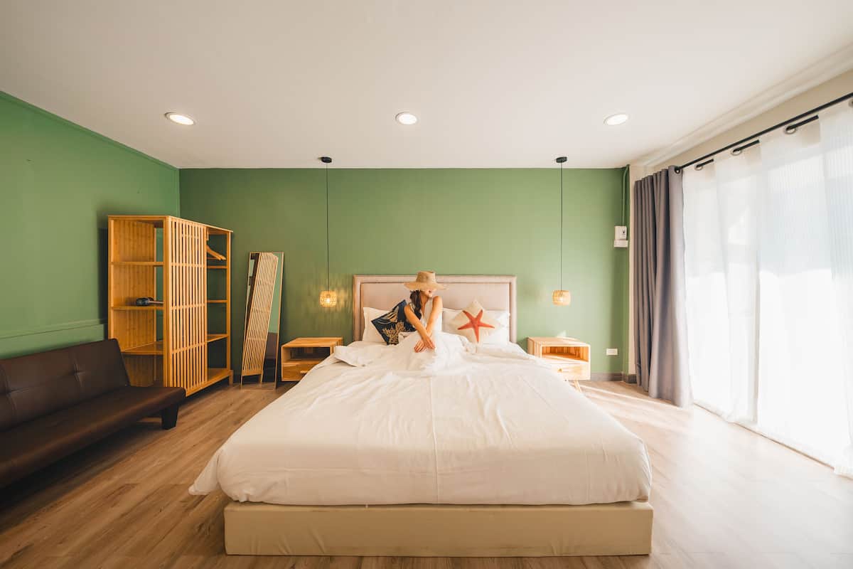 Uma cama grande com lençóis brancos, um guarda-roupa de madeira, um banco e uma mesa de cabeceira com abajur.