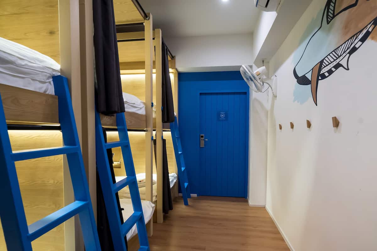 Um corredor com beliches, uma porta azul e um ventilador fixado na parede.