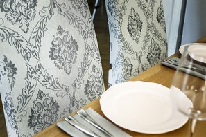 Uma mesa de jantar posta com um prato branco, talheres e um copo. A mesa está sobre uma superfície de madeira.
