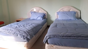 Duas camas de solteiro com cabeceiras, um pequeno criado-mudo de madeira e uma cadeira vermelha.